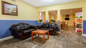 Um saguão de hotel com dois sofás de couro, uma mesa de centro de madeira, um quadro emoldurado na parede, uma área de refeições com cadeiras de madeira e um aviso anunciando o café da manhã.