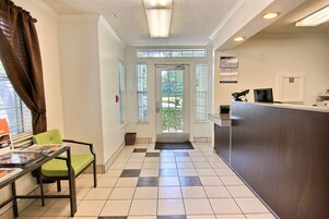 Uma área de recepção com balcão de check-in, uma cadeira verde, uma mesa com revistas e vista pela porta.