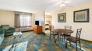 Quarto de hotel com área de estar, mesa de jantar com quatro cadeiras, televisão, pequena mesa com tampo de vidro, sofá, ventilador de teto e duas obras de arte emolduradas na parede.