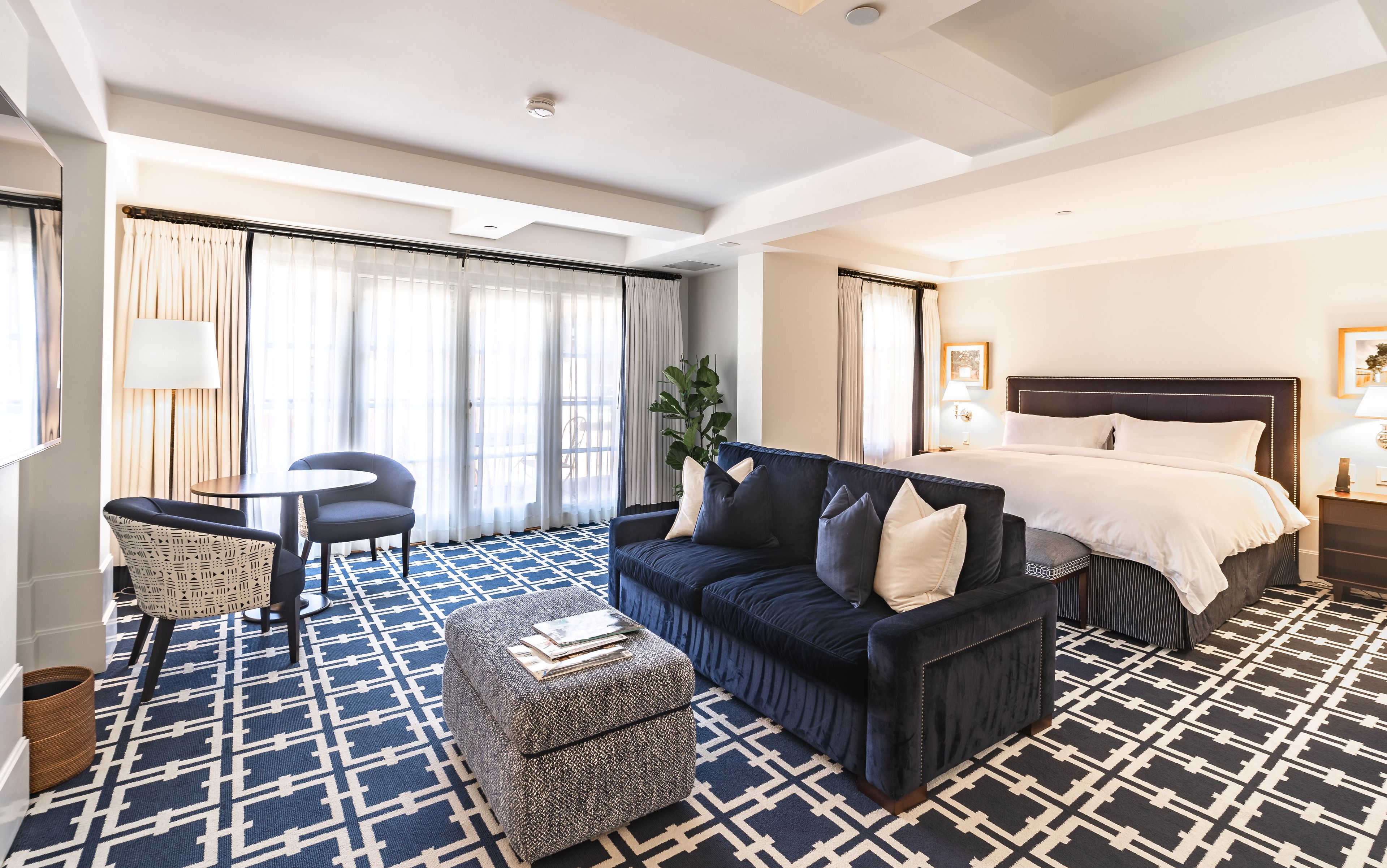 Quarto de hotel com uma cama grande, um sofá azul escuro, uma mesa redonda pequena e um piso estampado.