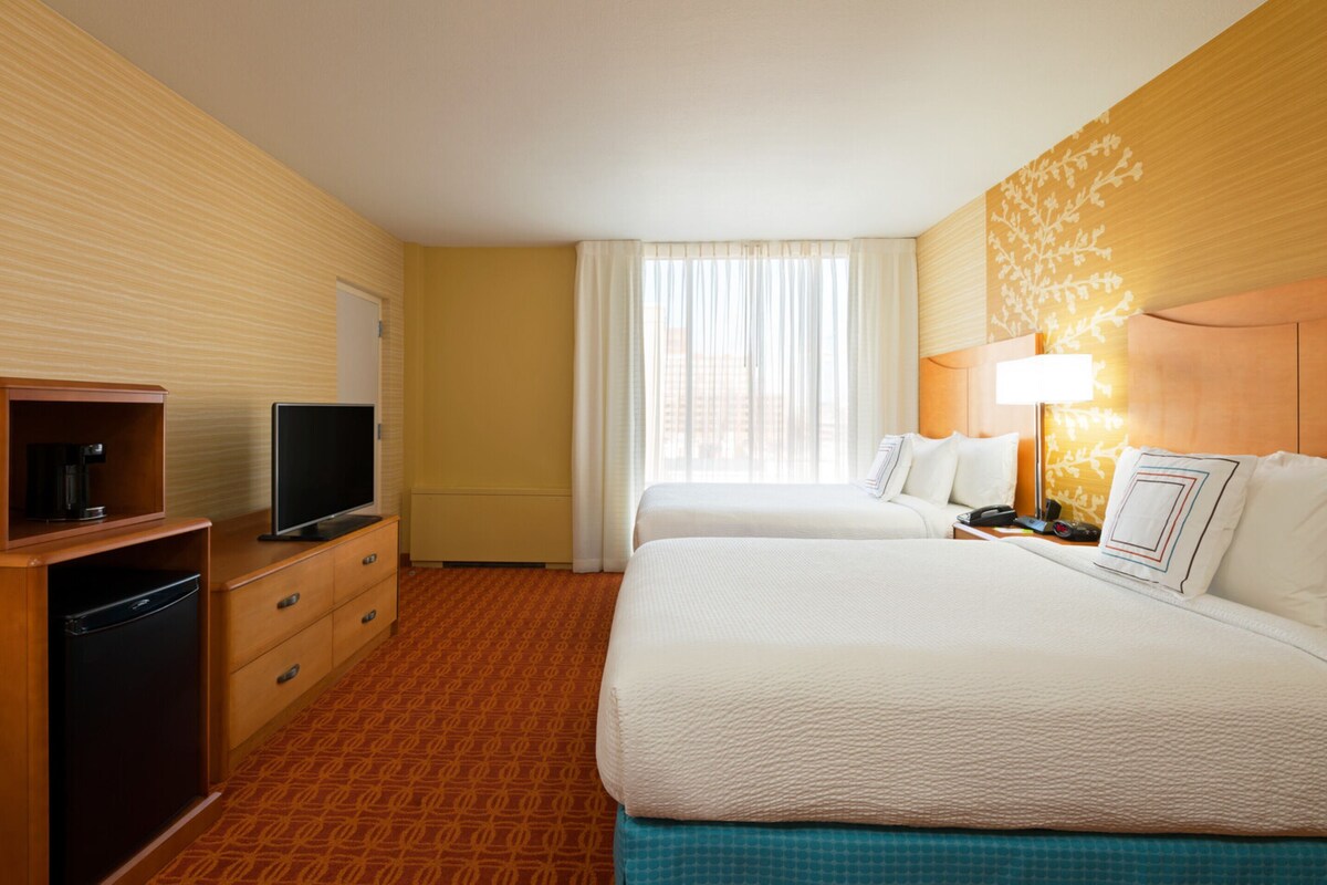 Quarto de hotel com duas camas, televisão, escrivaninha com computador e janela com cortinas.