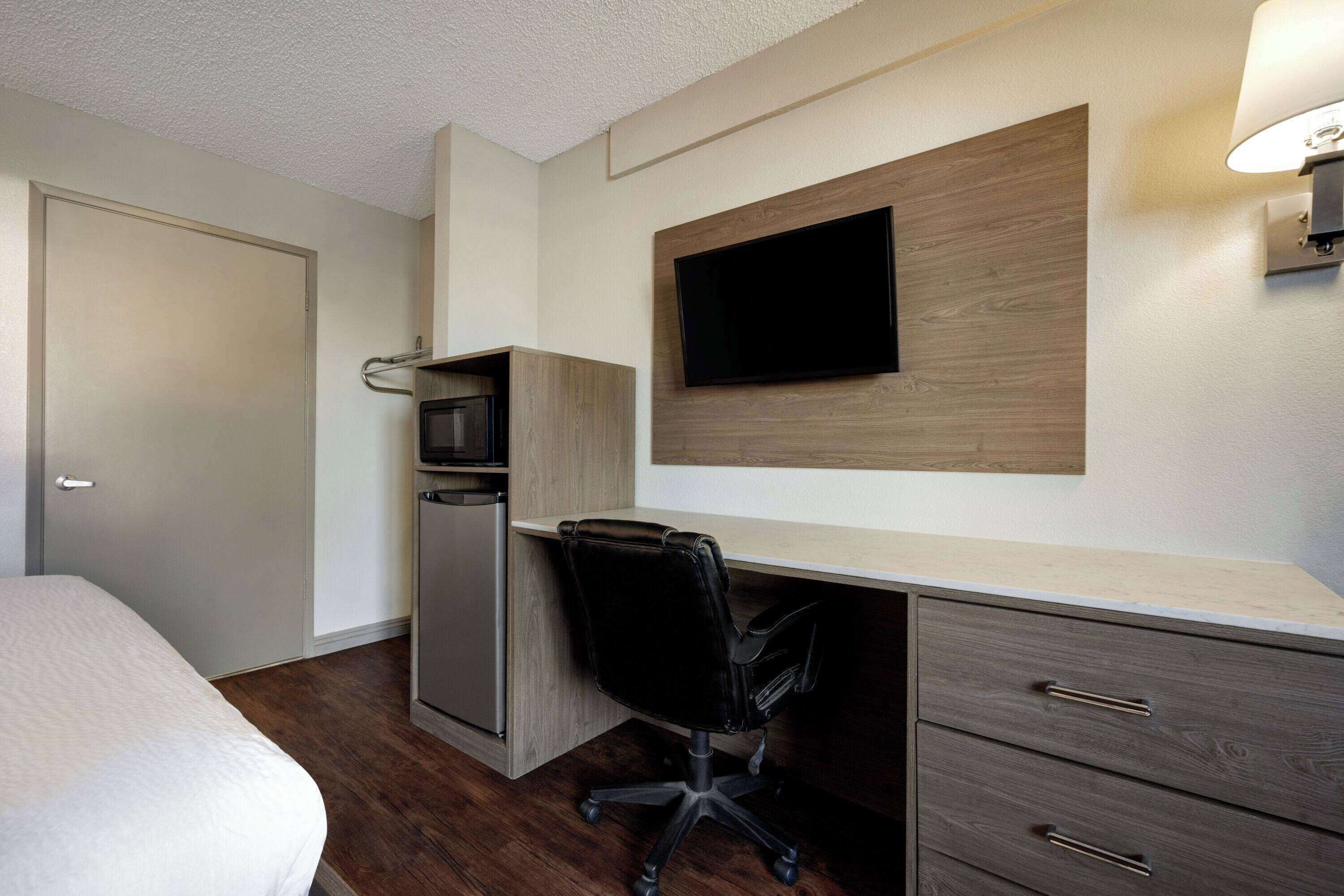 Quarto de hotel com cama, escrivaninha com cadeira, TV fixada na parede, micro-ondas e um pequeno frigorífico.