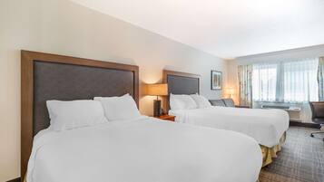 Quarto de hotel com duas camas, uma escrivaninha, uma cadeira, uma luminária e uma janela com cortinas.