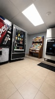 Máquina de vendas automática, expositor de snacks e dispensador de bebidas numa sala limpa e revestida a azulejos.