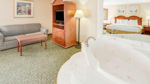 Quarto de hotel com sofá, armário de madeira com televisão, luminária de chão, banheira grande e cama com duas mesas de cabeceira.