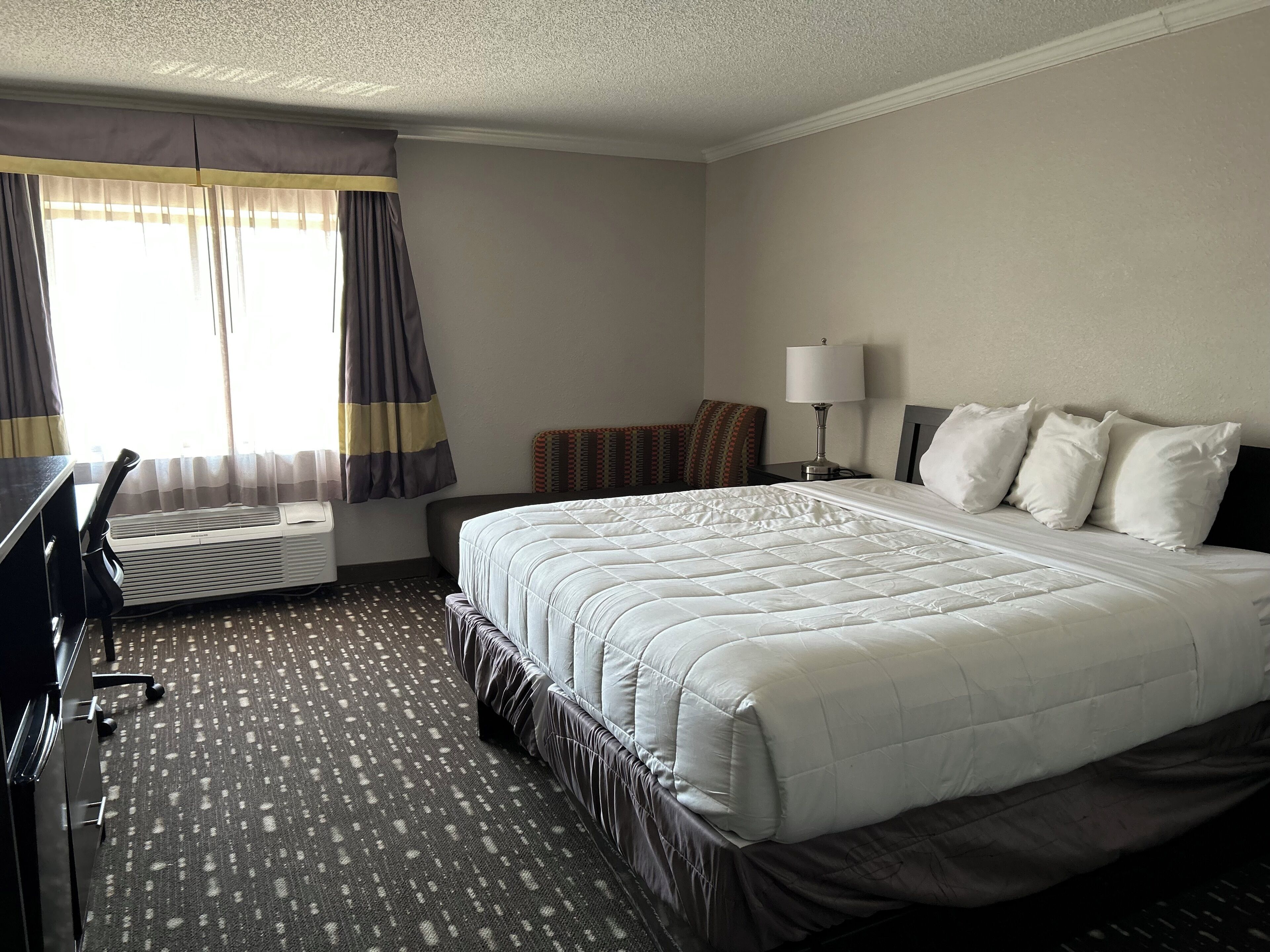 Quarto de hotel com uma cama grande, uma escrivaninha, uma cadeira, um abajur e uma janela com cortinas.