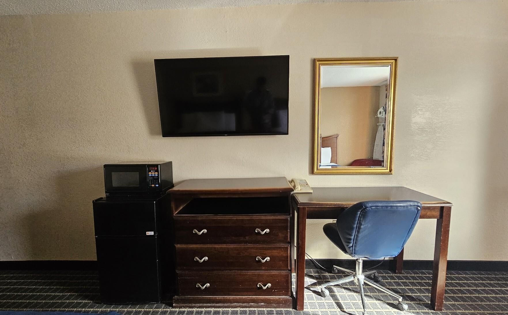 Quarto de hotel com escrivaninha de madeira, uma cadeira, televisão, micro-ondas e espelho.