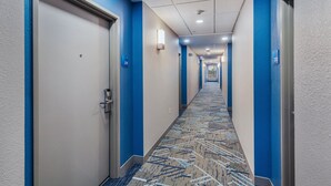 Corredor de hotel com paredes azuis e brancas, portas numeradas e um tapete estampado.