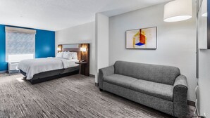 Quarto de hotel com uma cama, um sofá cinzento, um quadro e um abajur.