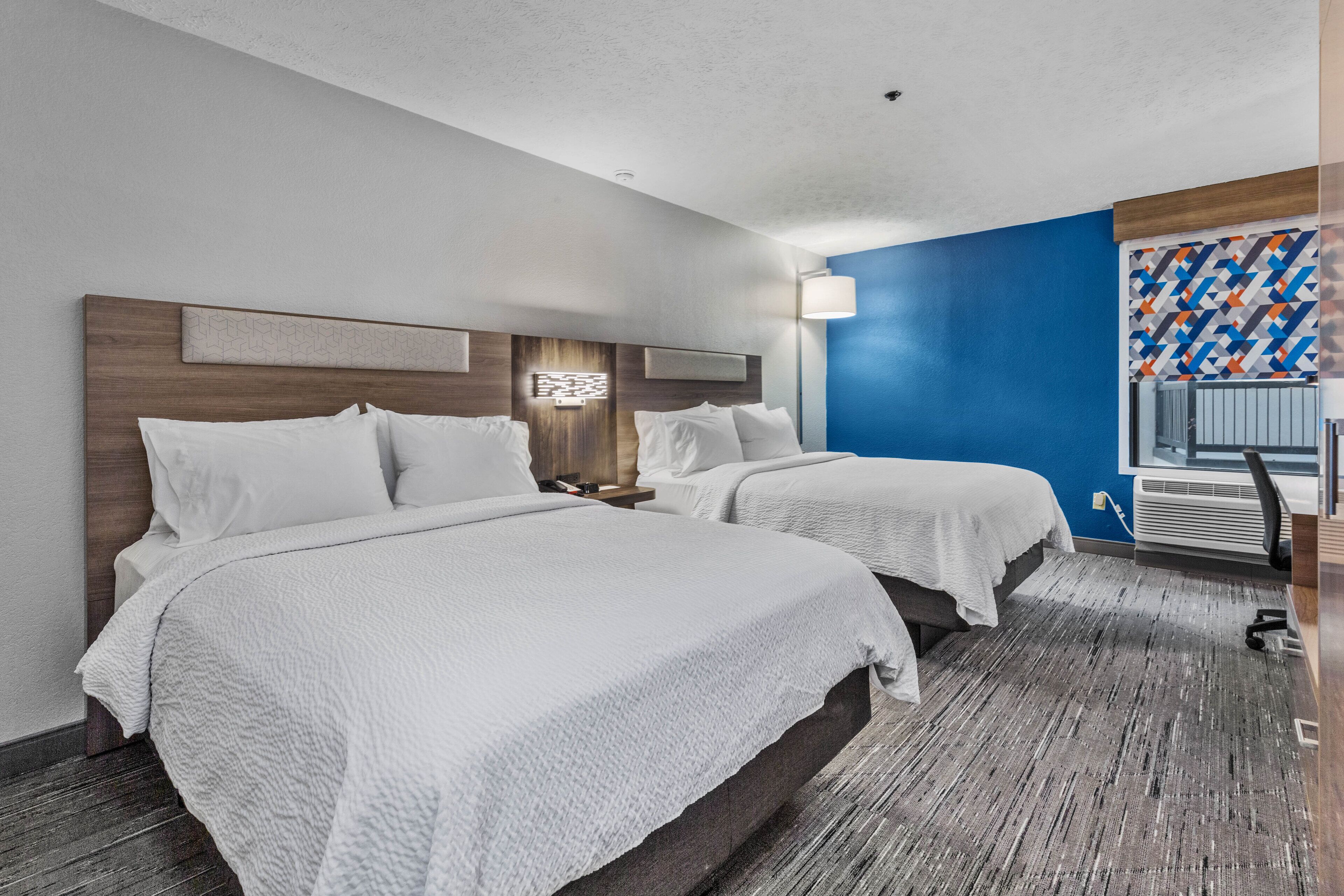 Quarto de hotel com duas camas, uma mesa de cabeceira, uma luminária na parede e uma janela com padrão geométrico.