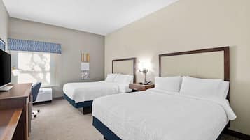 Quarto de hotel com duas camas, uma mesa, uma cadeira, televisão e janela com persianas.