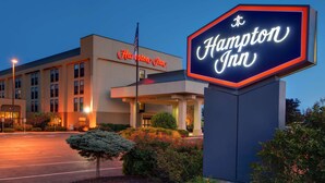 Um hotel Hampton Inn com uma entrada bem iluminada, uma placa grande e uma área ajardinada com arbustos e uma árvore pequena.