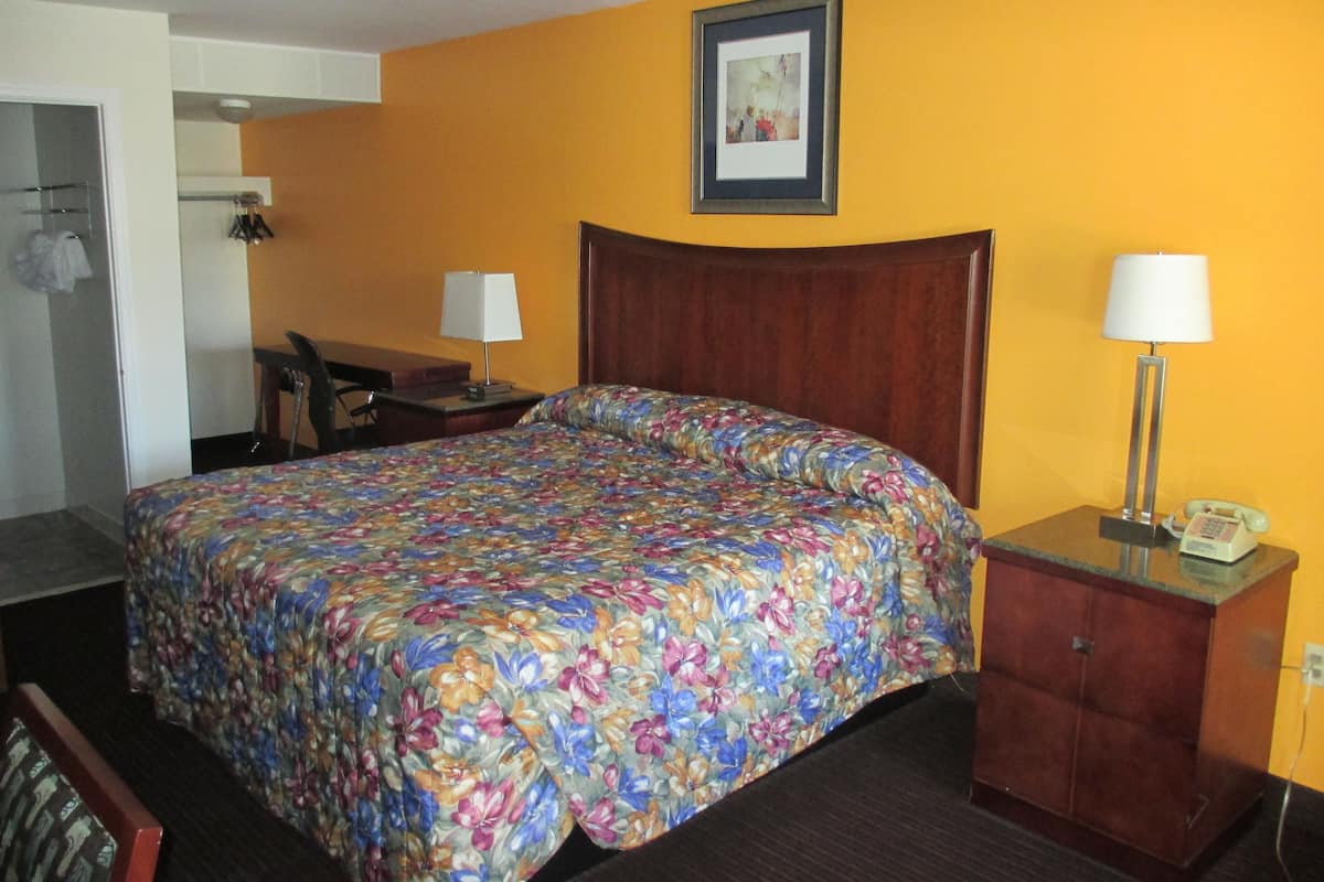 Quarto de hotel com cama, escrivaninha, cadeira, mesa de cabeceira com abajur e telefone.