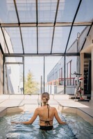 Pessoa em piscina coberta com paredes de vidro, com vista para um edifício e uma árvore lá fora.