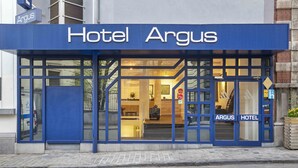 Entrada de um hotel com fachada azul, amplas janelas de vidro e uma placa com a inscrição 'Hotel Argus'.