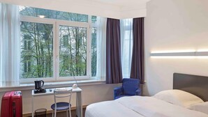 Quarto de hotel com cama, escrivaninha com cadeira, mala e vista para árvores pela janela.
