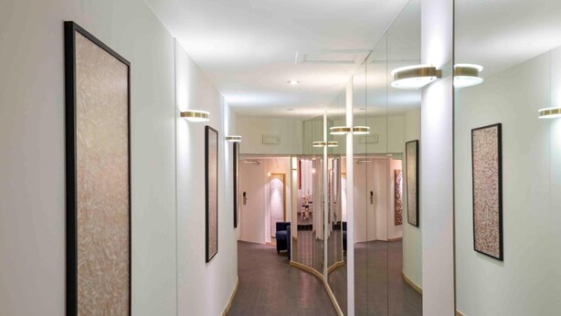 Um corredor com obras de arte emolduradas nas paredes, um espelho curvo refletindo o ambiente e iluminação embutida.