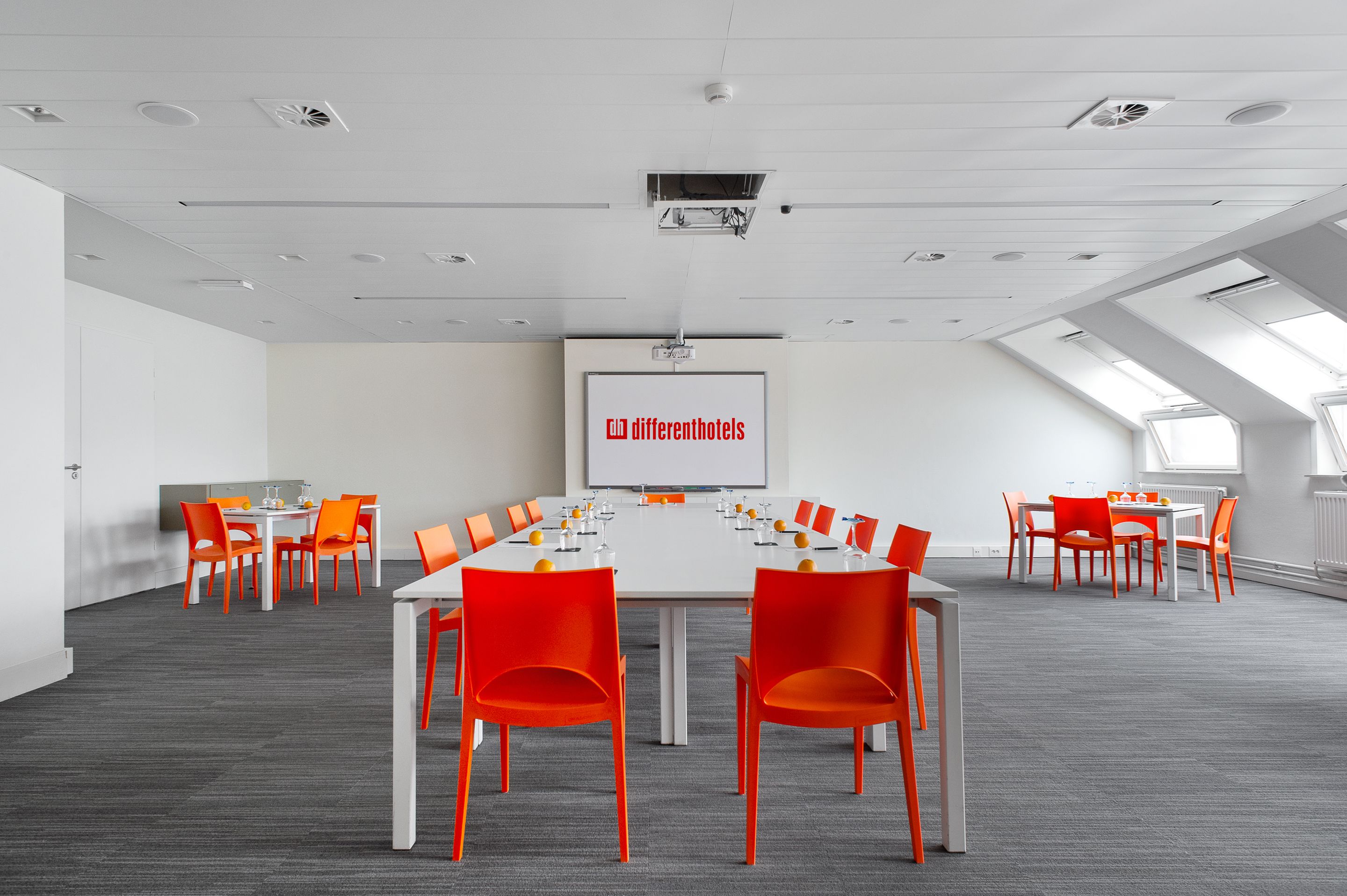 Uma sala de conferências com uma mesa grande e cadeiras laranja, um quadro branco com 'differenthotels' escrito e claraboias.