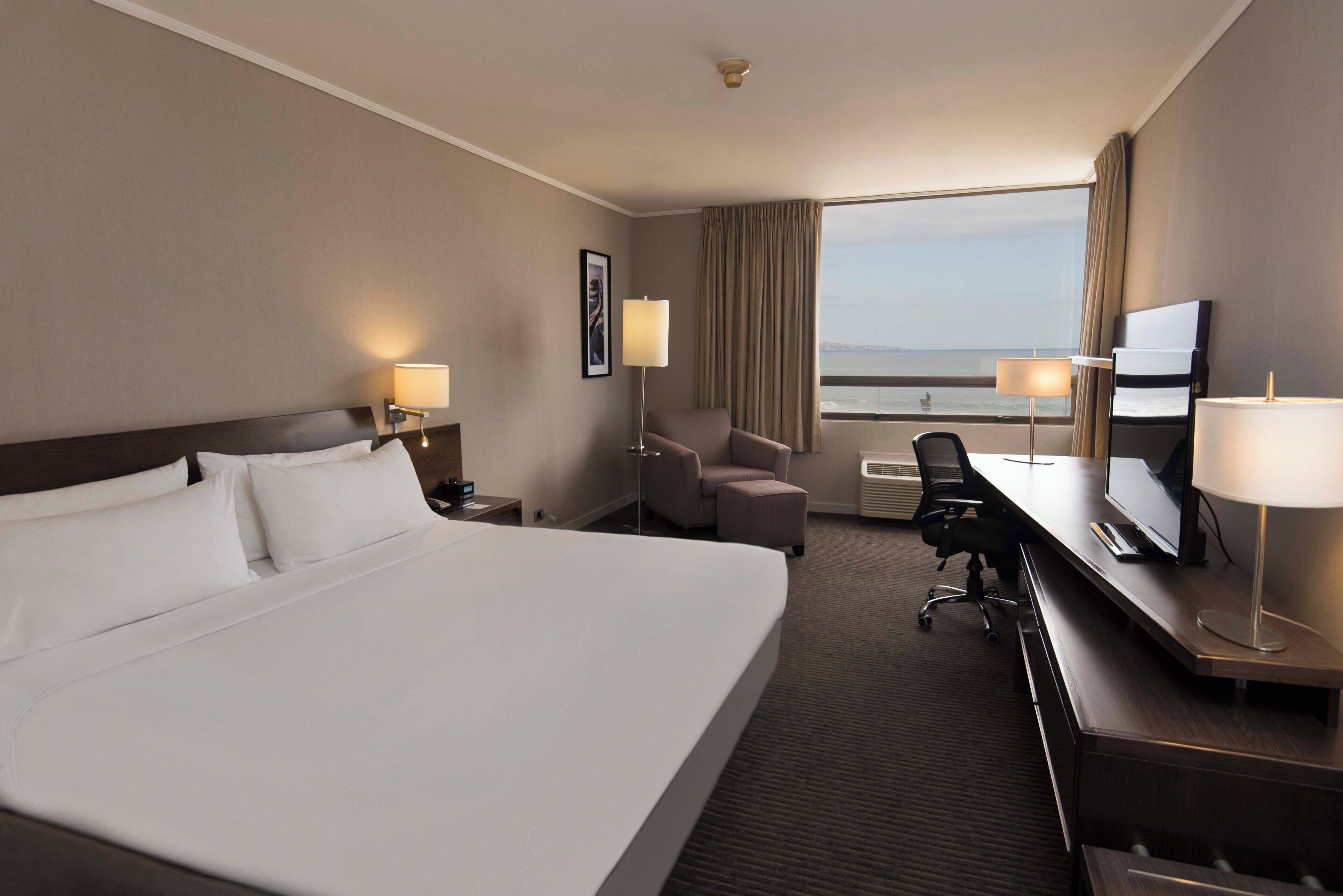Quarto de hotel com uma cama grande, uma escrivaninha com computador, uma cadeira, televisão e janela com vista.