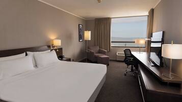 Quarto de hotel com uma cama grande, uma escrivaninha com computador, uma cadeira, televisão e janela com vista.