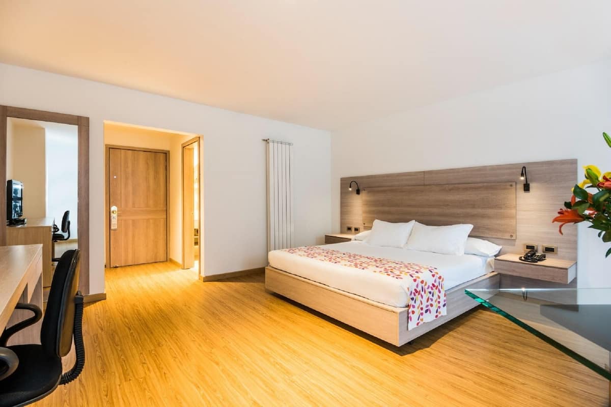 Quarto de hotel com chão de madeira, cama com roupa branca, escrivaninha com cadeira, mesa de cabeceira com abajur e espelho na parede.