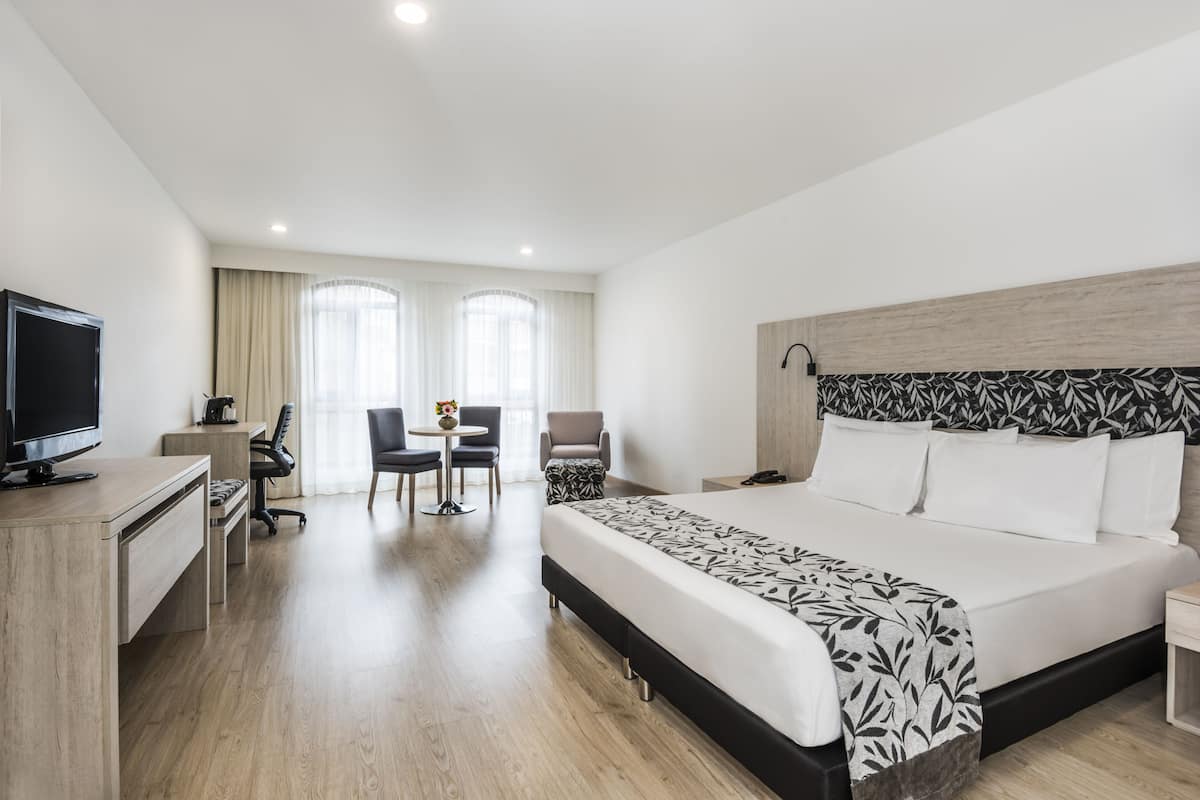 Quarto de hotel com uma cama grande, uma escrivaninha com computador, televisão e duas cadeiras.