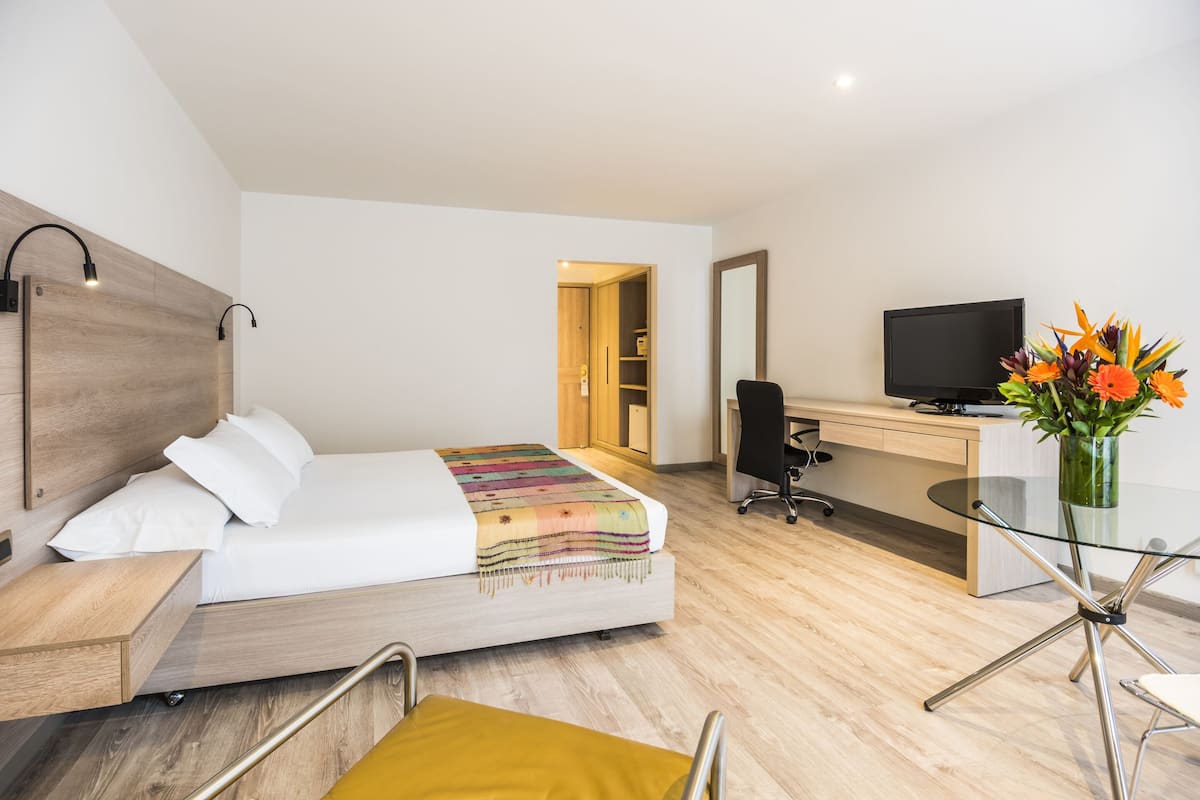 Quarto de hotel moderno com cama, escrivaninha, cadeira, televisão e uma mesa de vidro com um vaso de flores.