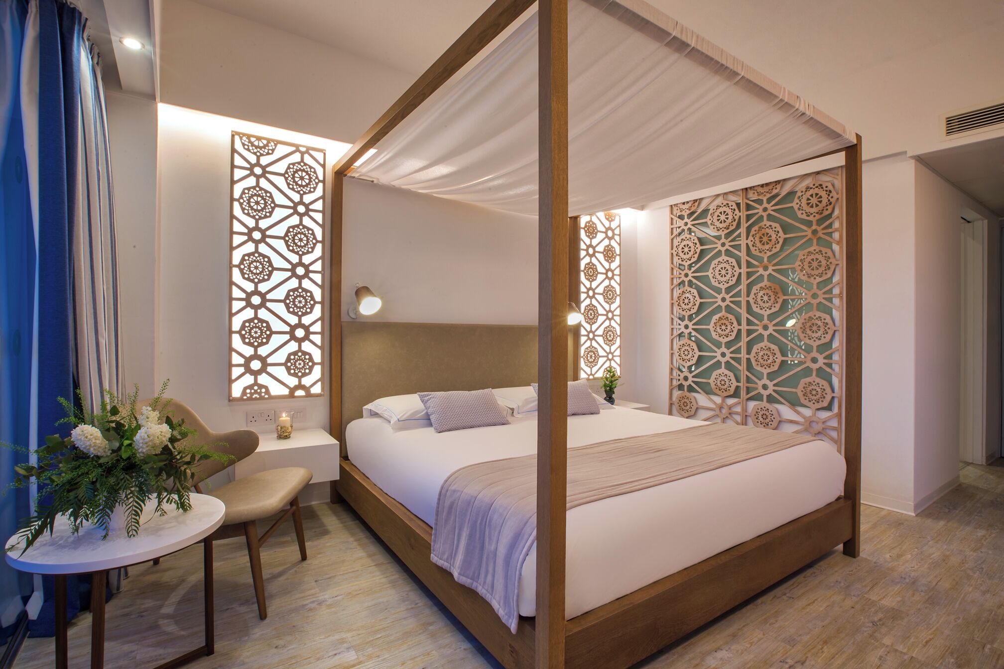 Uma cama com dossel e estrutura de madeira, roupa de cama branca, uma pequena mesa de cabeceira com abajur e um painel decorativo na parede com padrões geométricos.