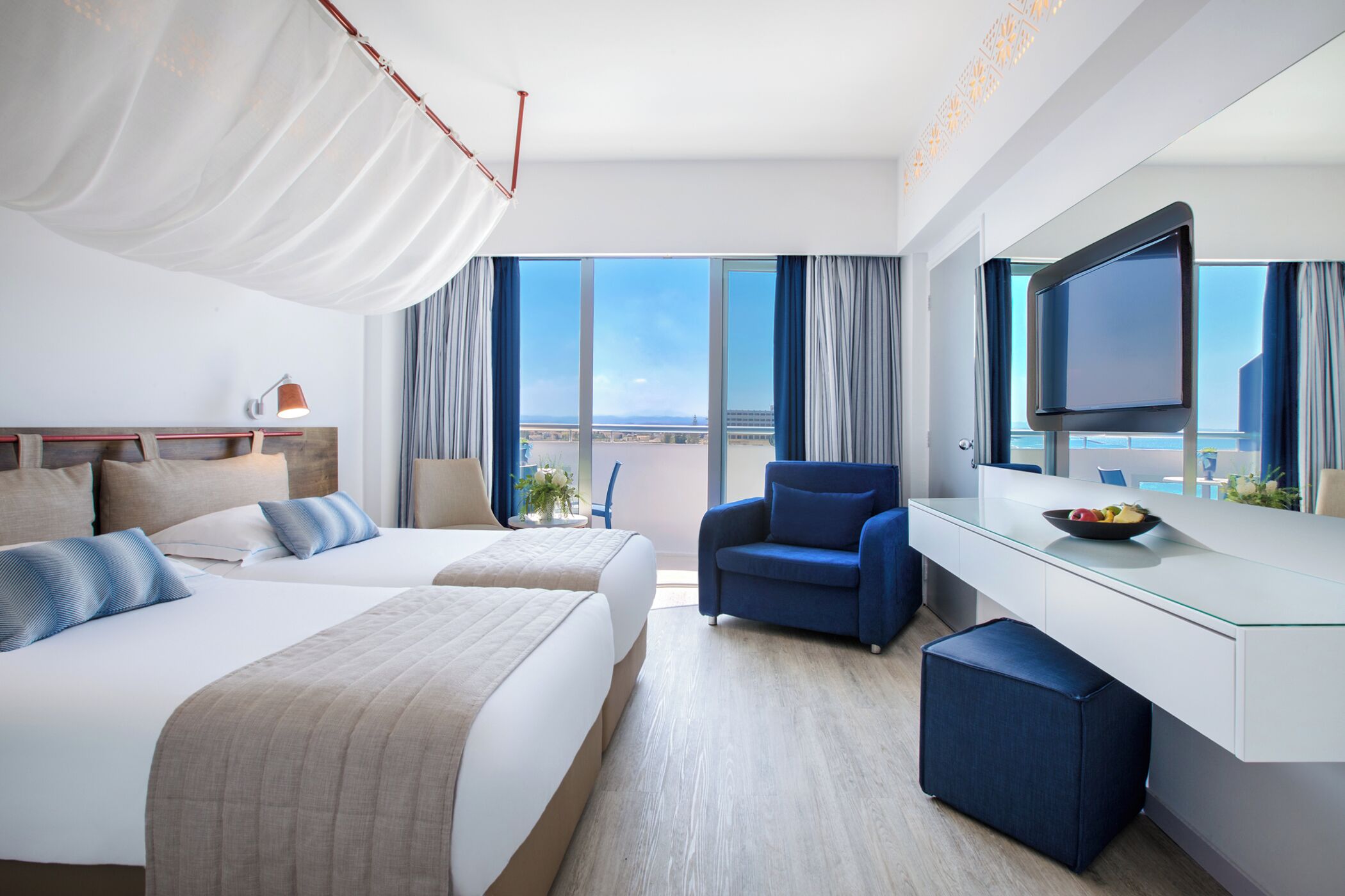 Quarto de hotel moderno com duas camas, uma escrivaninha com televisão e janelas amplas com vista para a cidade.