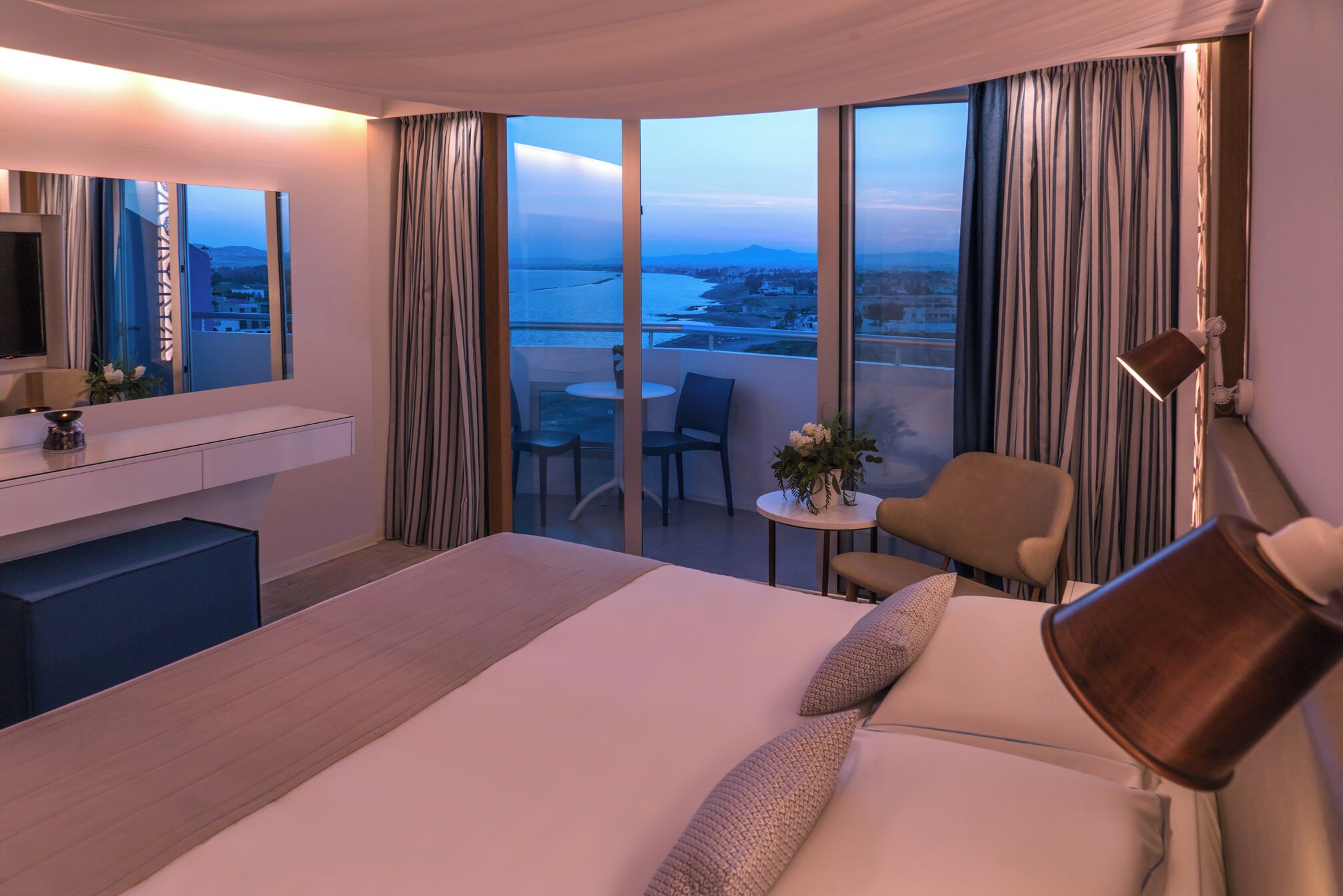 Quarto de hotel com uma cama grande, uma escrivaninha, uma cadeira, uma mesinha e vista para o mar.