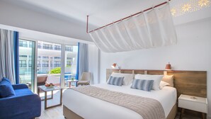 Quarto de hotel moderno com uma cama grande, um sofá azul, uma mesinha com frutas e vista da cidade através de amplas janelas.