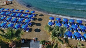 Vista aérea de uma praia com fileiras de guarda-sóis azuis e cadeiras de praia brancas, palmeiras e uma vista clara do oceano.