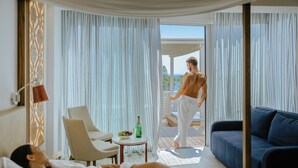Quarto de hotel com uma cama, um sofá, uma mesa com uma garrafa e frutas, e vista para o exterior através das cortinas.