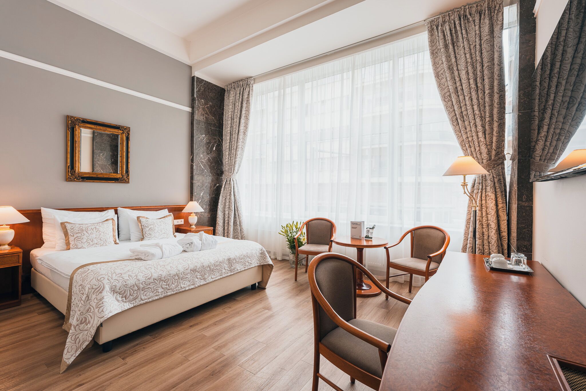 Quarto de hotel com cama, mesas de cabeceira, escrivaninha, cadeiras e uma janela grande com cortinas.