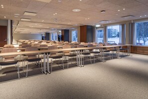 Uma sala de conferências com fileiras de mesas e cadeiras, grandes janelas com vista para árvores e edifícios, e um teto com iluminação embutida.