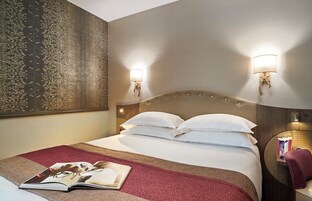 Quarto de hotel com uma cama grande, duas luminárias fixadas na parede, um cobertor vermelho e uma revista sobre a cama.