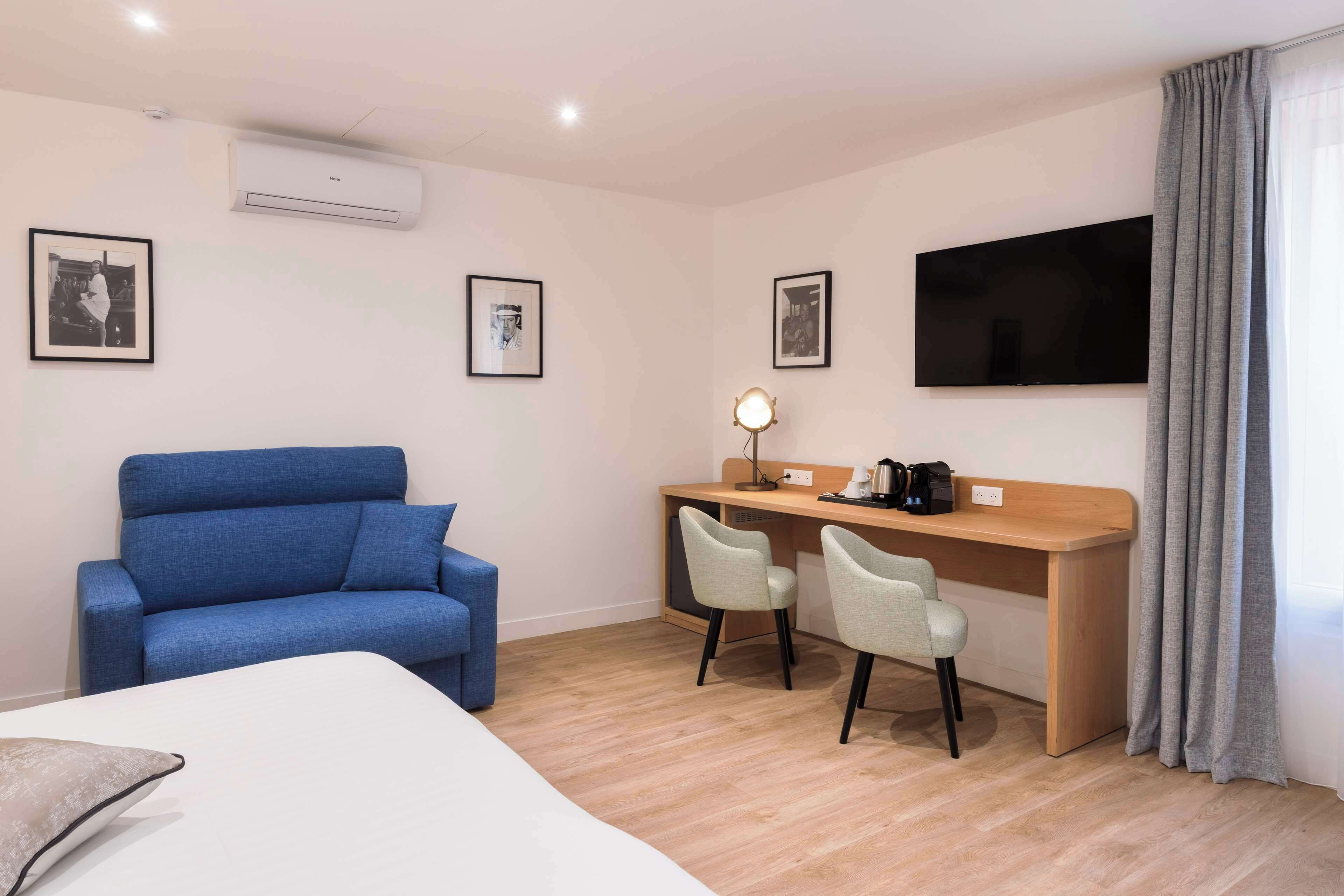 Quarto de hotel com cama, sofá azul, escrivaninha com duas cadeiras, televisão de tela plana e quadros nas paredes.