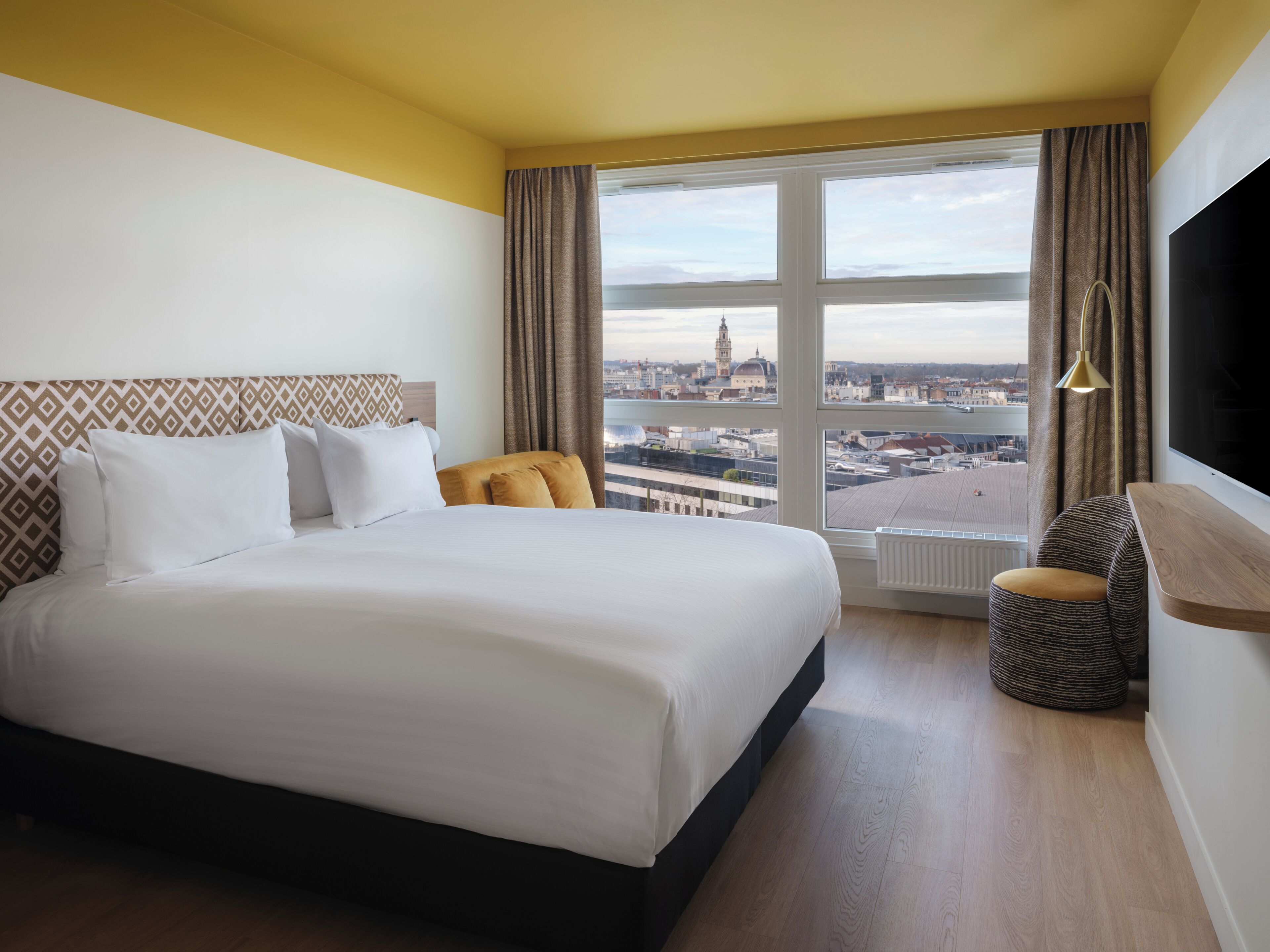 Quarto de hotel com uma cama grande, vista para a cidade, televisão e um pequeno espaço para sentar.