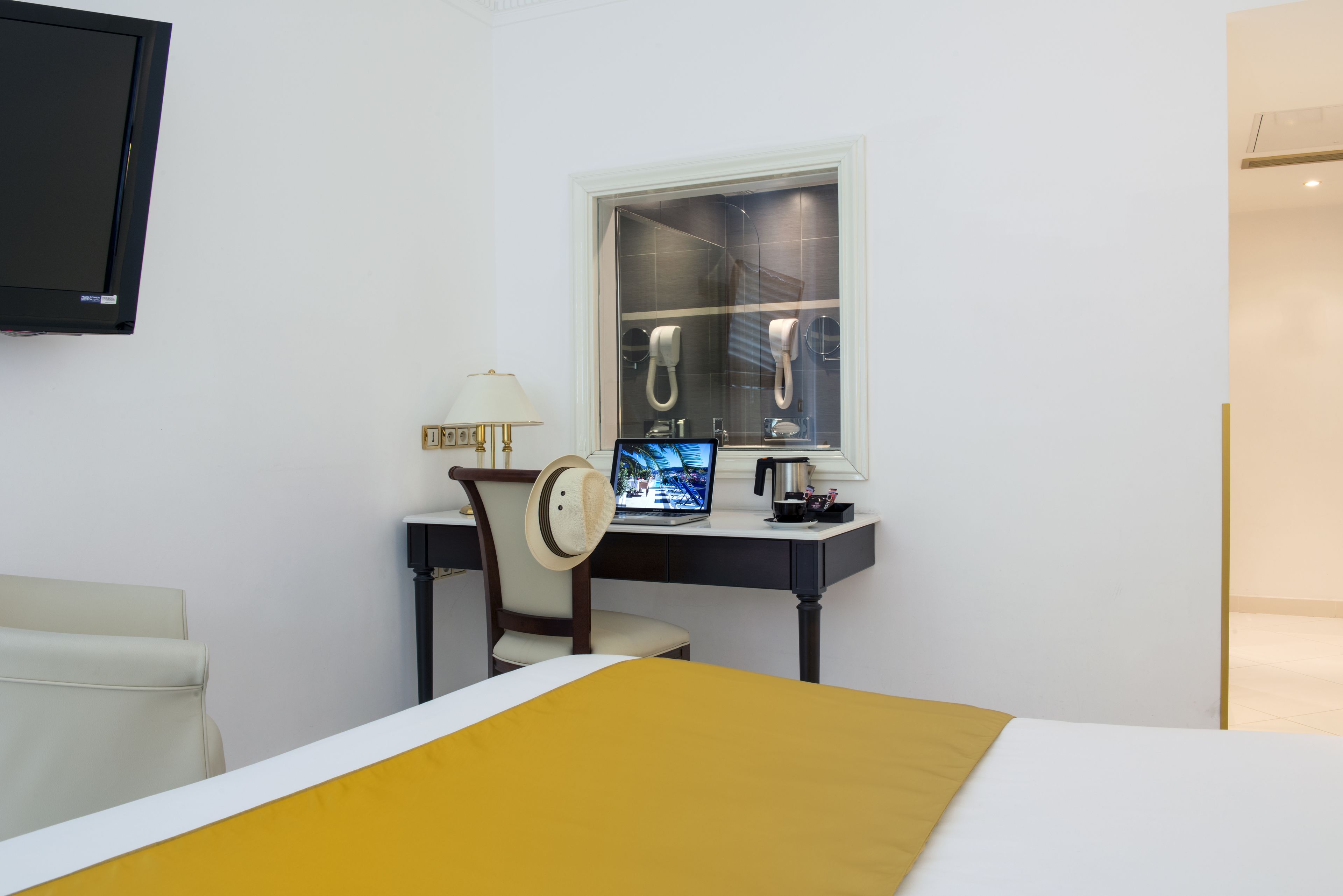 Quarto de hotel com cama, escrivaninha com laptop, cadeira, abajur e telefone.