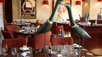 Interior de restaurante com mesas postas, cadeiras vermelhas estofadas, um espelho grande e luminárias vermelhas fixadas na parede.