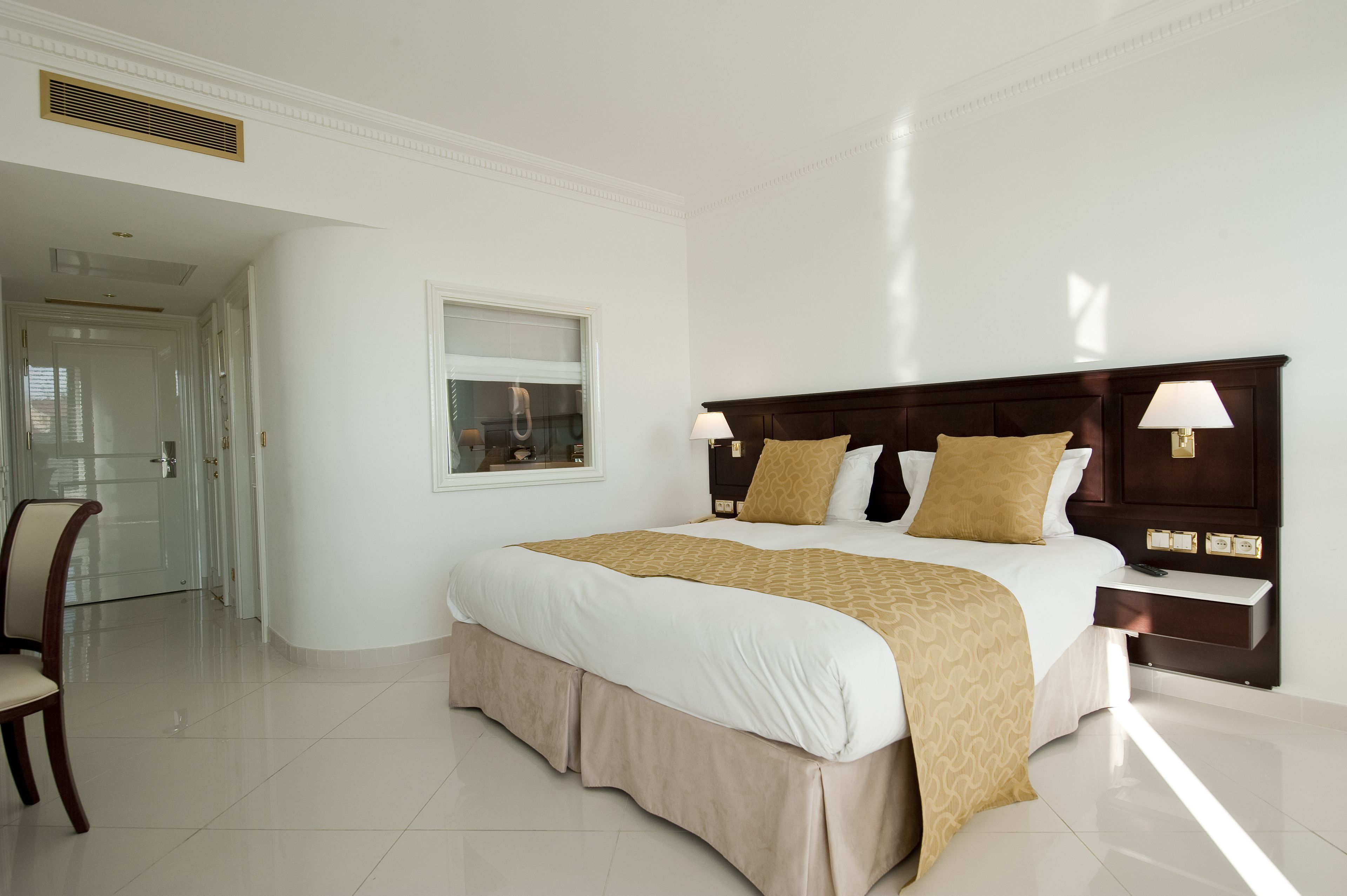 Quarto de hotel com uma cama grande, mesa de cabeceira com luminárias, um espelho e uma cadeira.