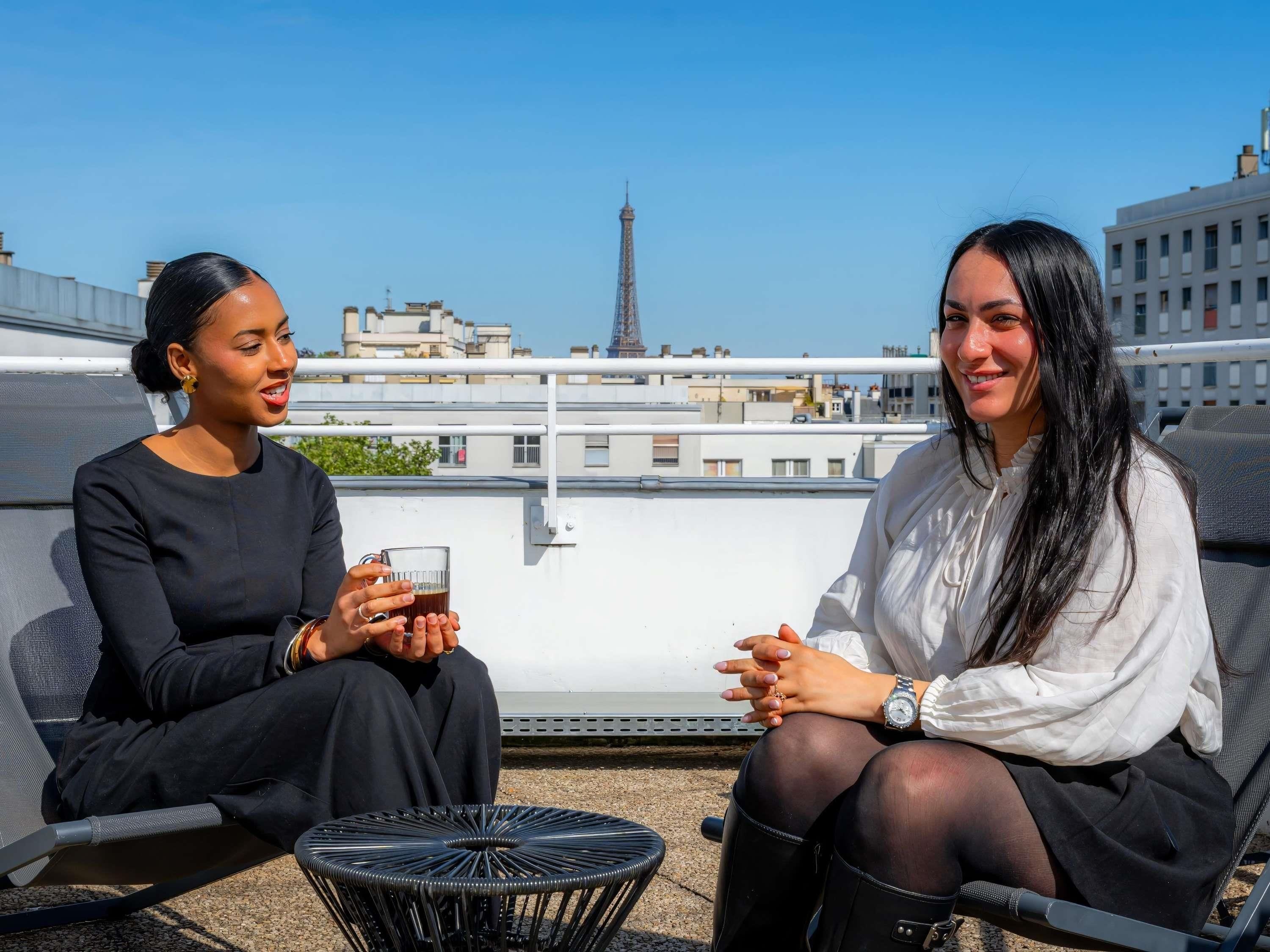 Duas pessoas sentadas em um terraço no telhado, com vista para uma paisagem urbana e uma torre ao fundo.