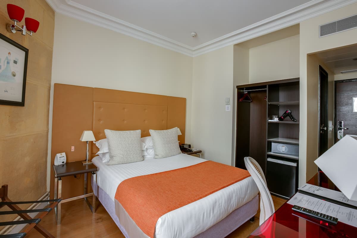 Quarto de hotel com cama, mesa de cabeceira, abajur e escrivaninha com cadeira.