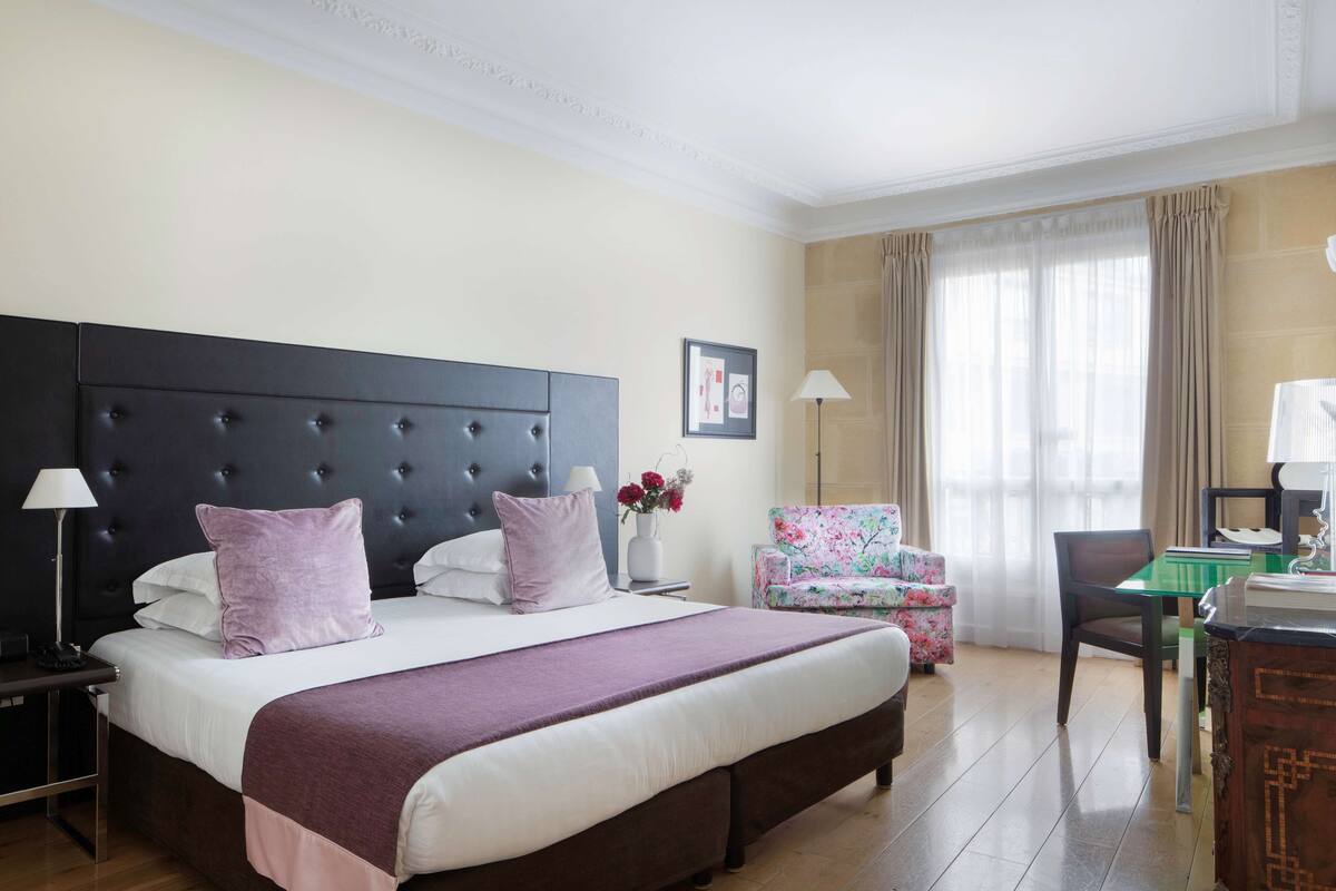 Quarto de hotel com uma cama grande, uma mesa, uma cadeira e uma janela com cortinas.