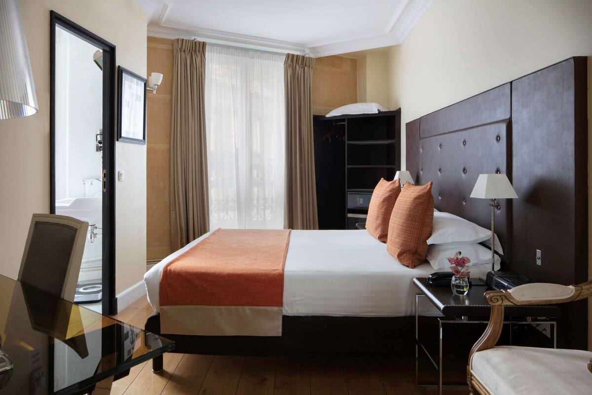 Quarto de hotel com uma cama grande, uma escrivaninha, uma cadeira, um guarda-roupa e uma janela com cortinas.