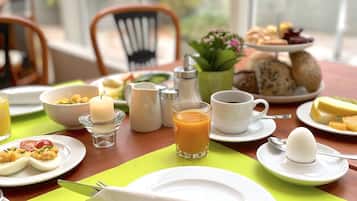 Uma mesa de café da manhã com diversos alimentos, um copo de suco de laranja e vista pela janela.