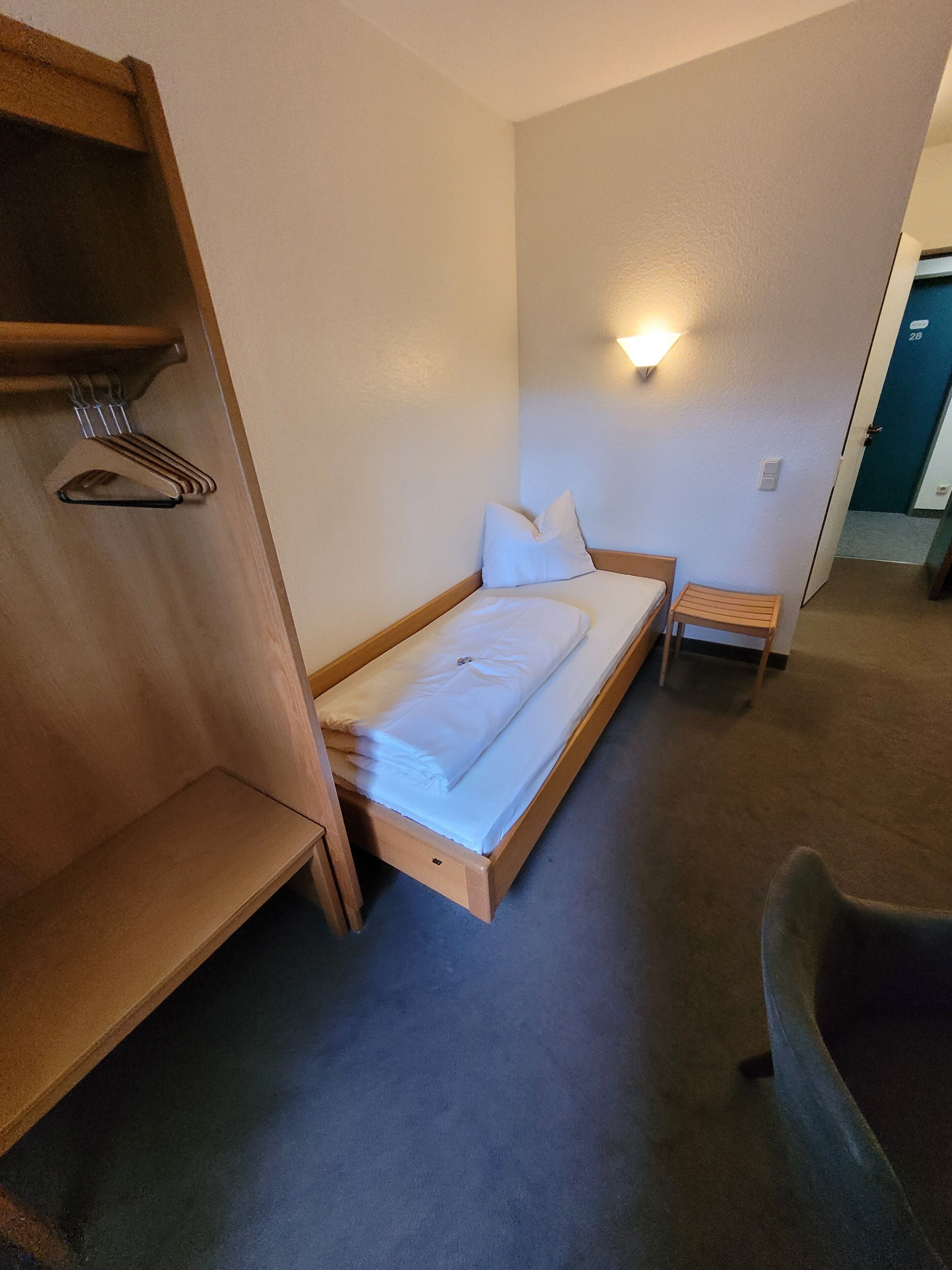 Quarto pequeno com uma cama de solteiro, um banco de madeira, uma luminária na parede e uma porta que dá para outro quarto.