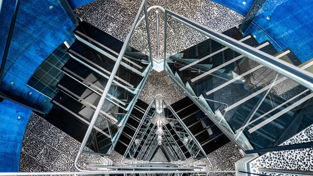 Estrutura arquitetônica moderna com um eixo central de elevador de vidro, cercado por grades de metal e um piso estampado.
