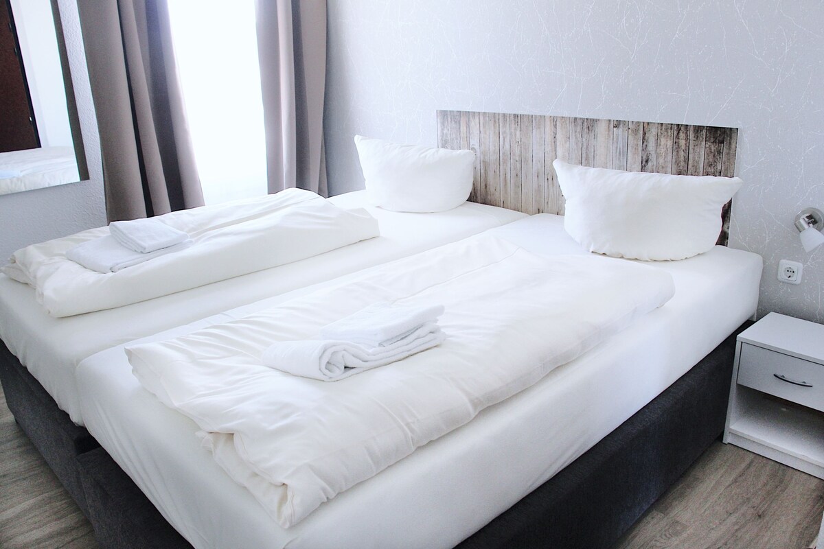 Uma cama de casal com lençóis e almofadas brancos, cabeceira de madeira, mesa de cabeceira com abajur e uma janela com cortinas.