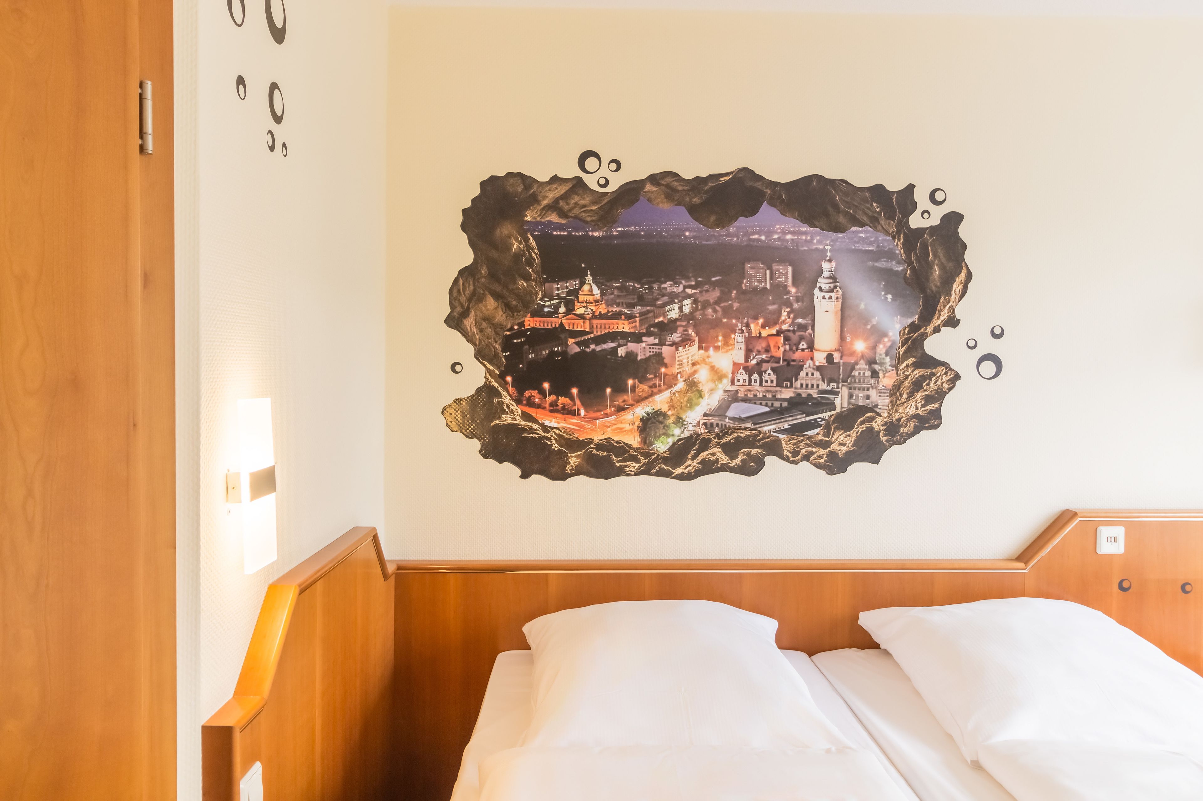 Quarto de hotel com cama, cabeceira de madeira, luminária fixada na parede e um mural com vista para a cidade.